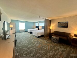 Quarto de hotel com duas camas, um sofá, uma escrivaninha com computador e uma televisão.