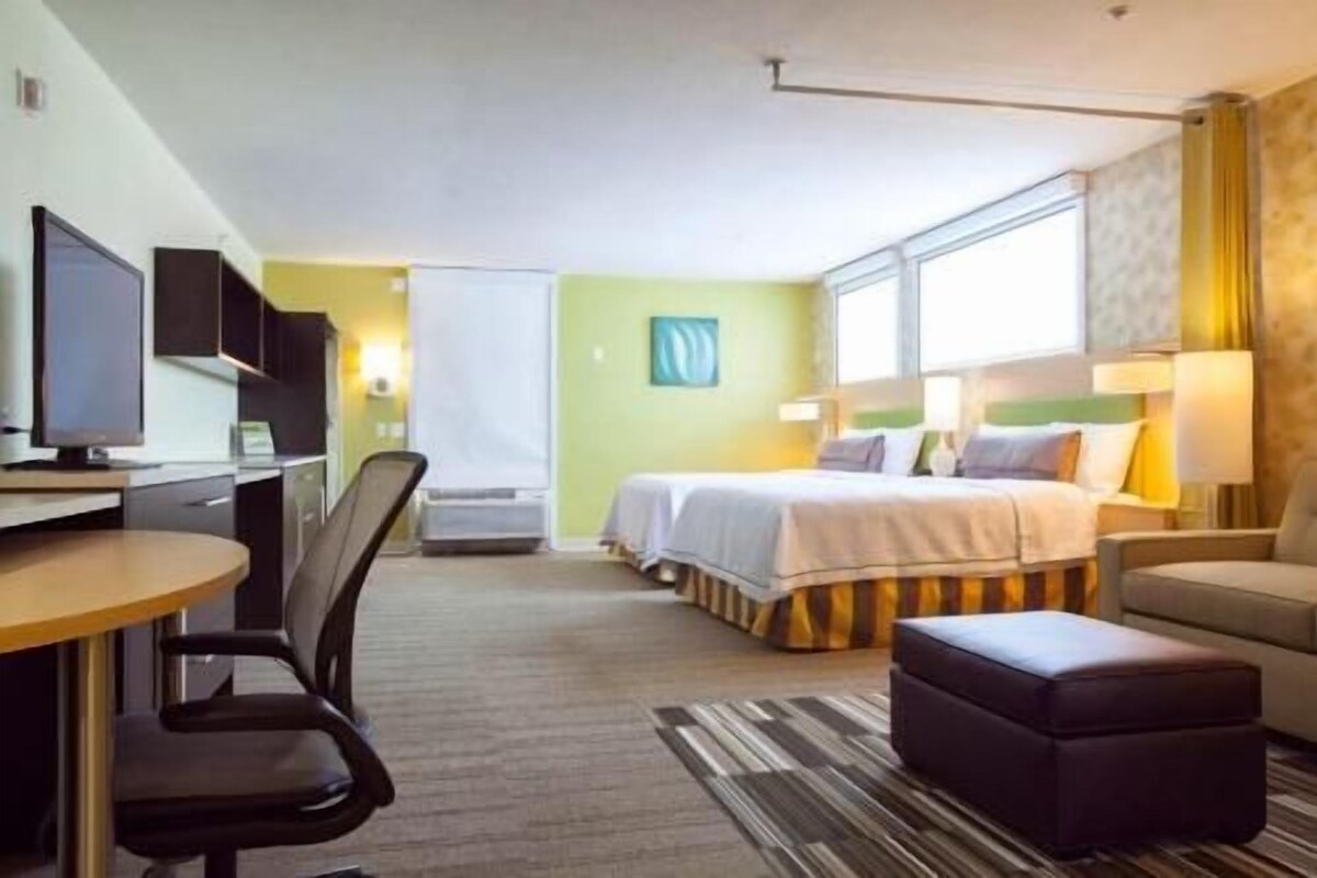 Quarto de hotel com uma cama, uma escrivaninha com computador, uma cadeira e uma janela.