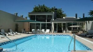 Piscina com espreguiçadeiras e guarda-sóis, cercada por um edifício com janelas amplas e um terraço coberto.