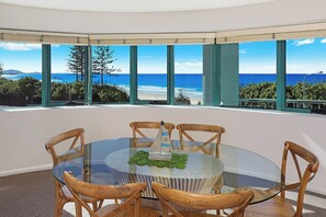 Mesa de vidro redonda com cadeiras de madeira em um quarto com grandes janelas com vista para uma praia.
