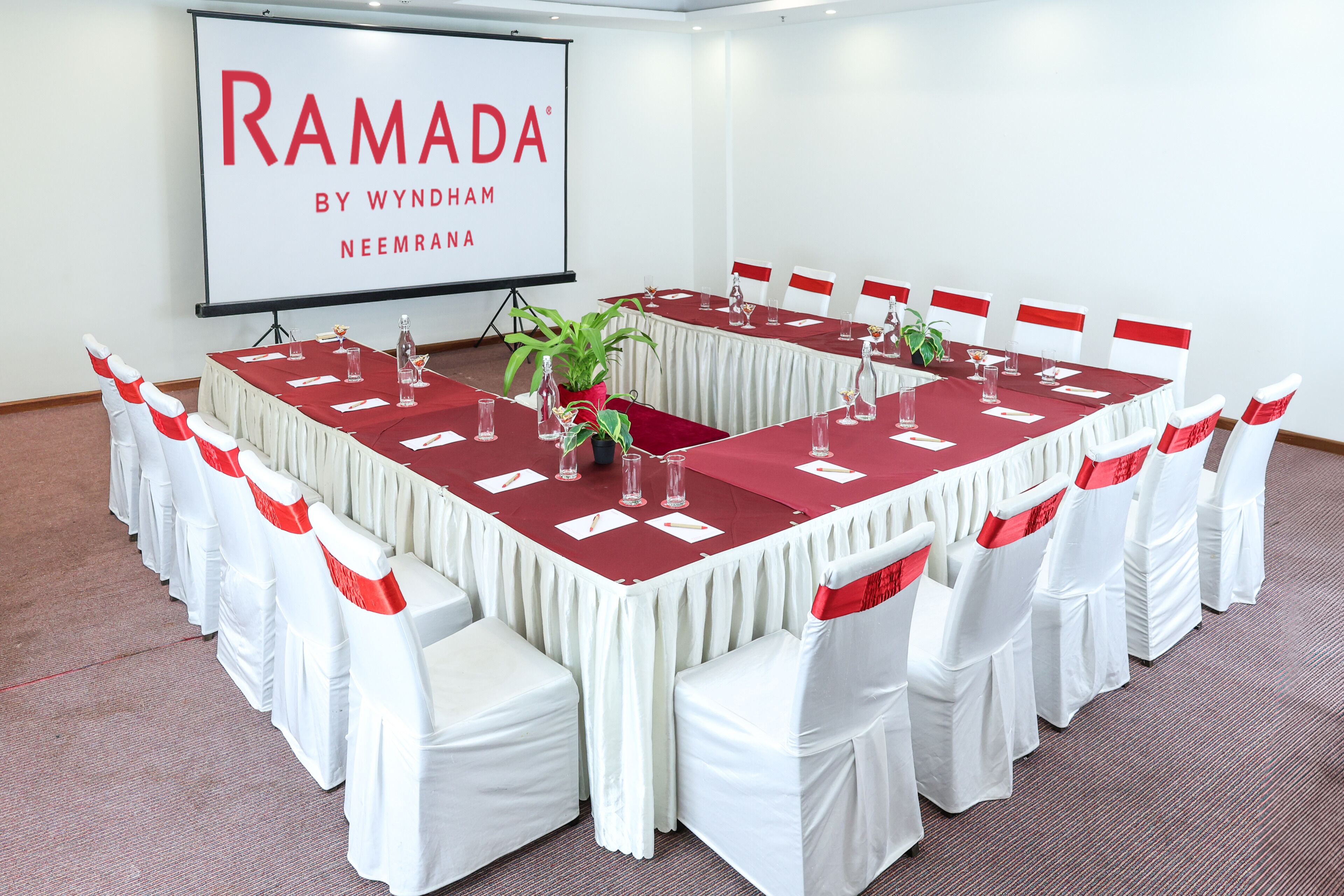Uma sala de conferências com uma mesa grande, cadeiras brancas com fitas vermelhas e uma tela de projetor exibindo 'RAMADA BY WYNDHAM NEEMRANA'.