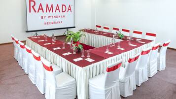 Uma sala de conferências com uma mesa grande, cadeiras brancas com fitas vermelhas e uma tela de projetor exibindo 'RAMADA BY WYNDHAM NEEMRANA'.