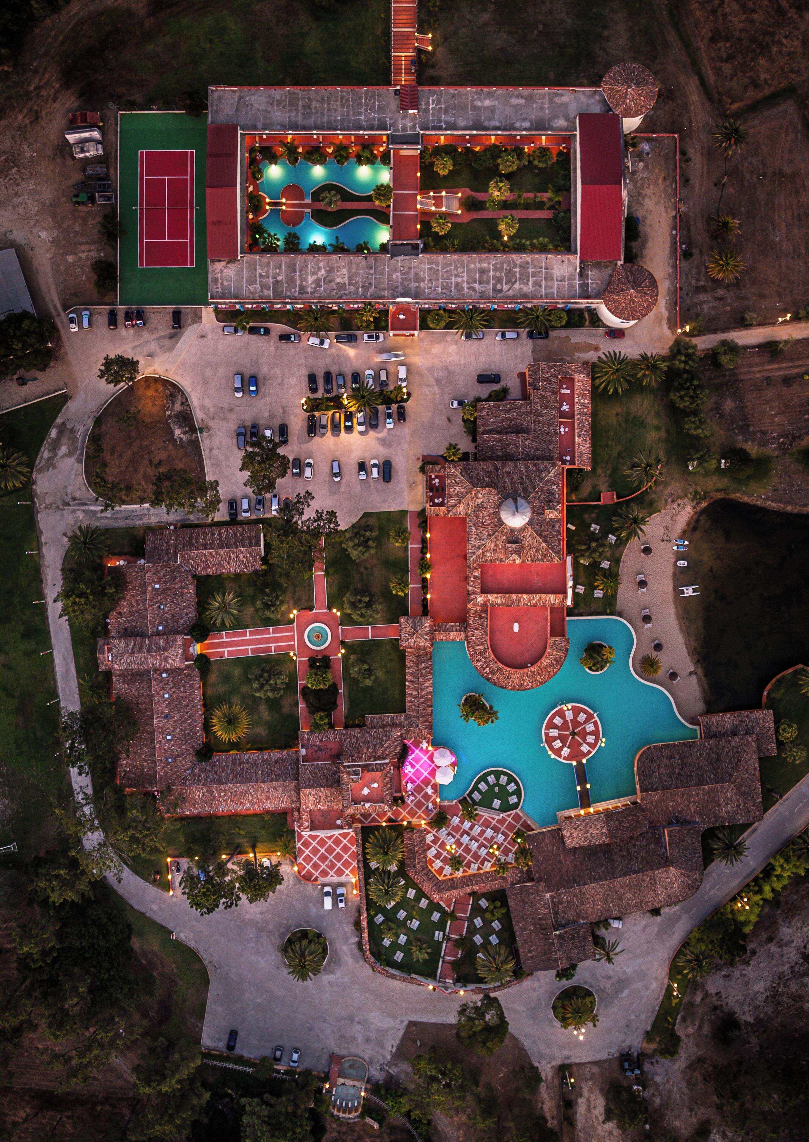 Vista aérea de um resort com piscina, quadra de tênis e várias áreas de estacionamento.