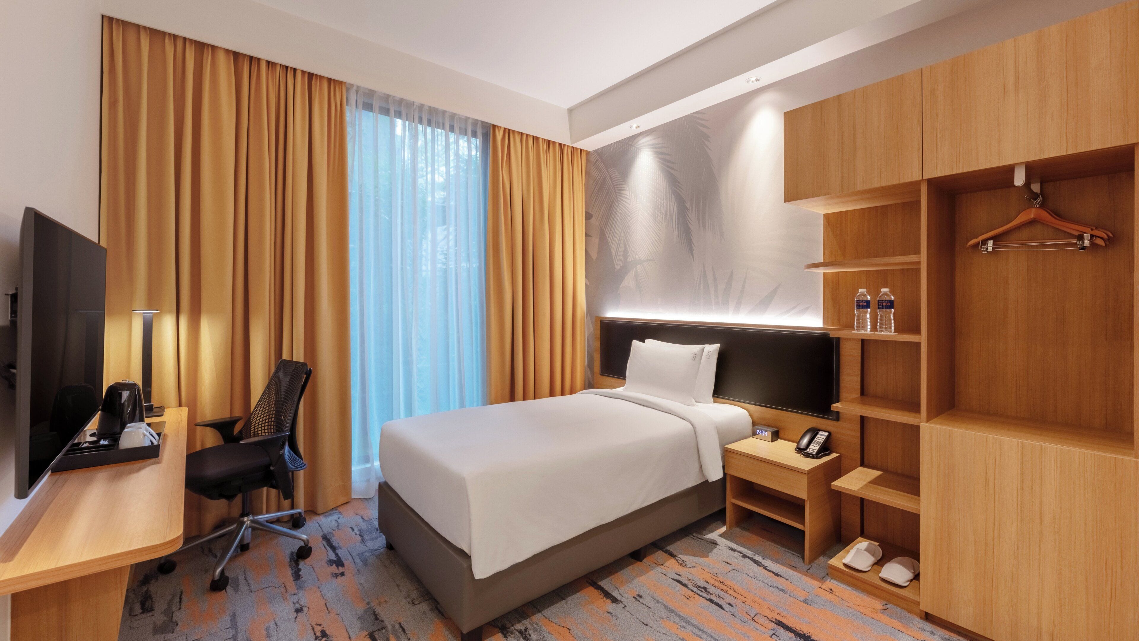 Quarto de hotel com cama, escrivaninha, cadeira, televisão, mesa de cabeceira com telefone e abajur, guarda-roupa e janela com cortinas.