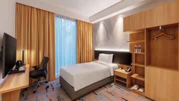 Quarto de hotel com cama, escrivaninha, cadeira, televisão, mesa de cabeceira com telefone e abajur, guarda-roupa e janela com cortinas.