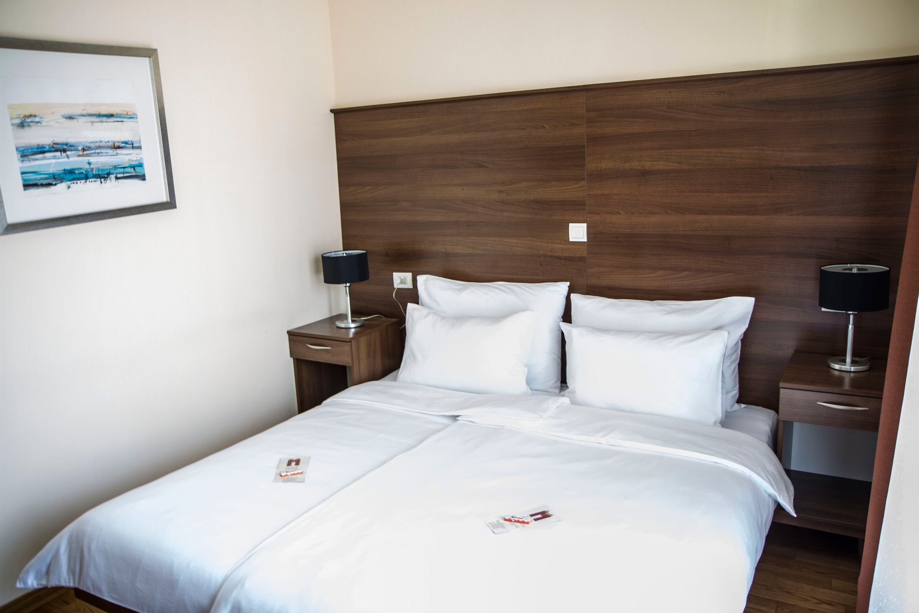 Quarto de hotel com cabeceira de madeira, roupa de cama branca, duas mesas de cabeceira com abajures e um quadro emoldurado na parede.