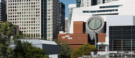 Um edifício moderno com um elemento de design circular marcante, rodeado por um espelho d'água e arranha-céus urbanos.