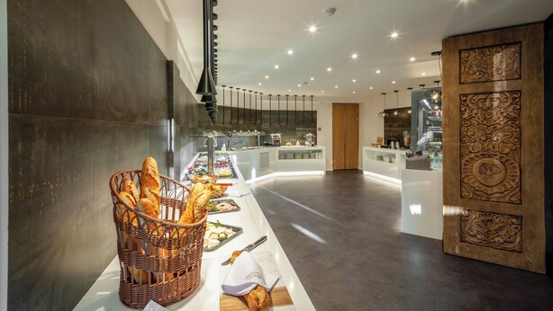 Um lobby moderno de hotel com área de buffet, com pães e doces, um balcão com diversas opções de comida e um painel decorativo de madeira.