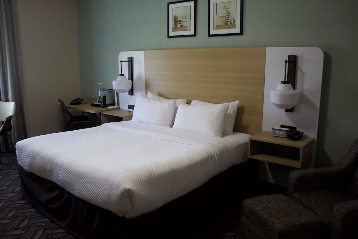 Quarto de hotel com uma cama grande, uma escrivaninha, uma cadeira, uma mesinha de cabeceira com abajur e duas quadros nas paredes.