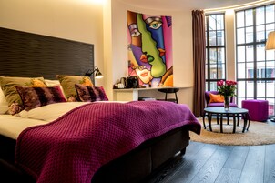 Um quarto moderno com uma cama grande, uma pintura abstrata vibrante, uma pequena mesa redonda e vista da cidade por grandes janelas.