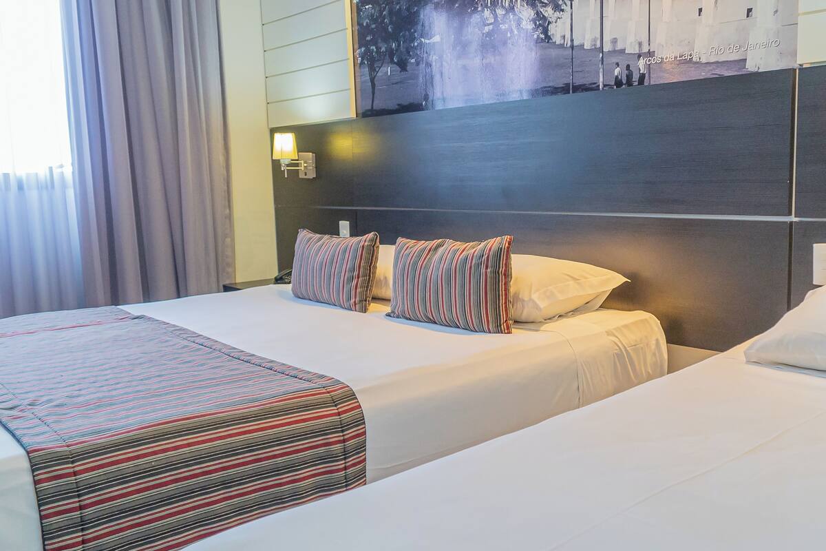 Quarto de hotel com duas camas, roupa de cama listrada, cabeceira com luz noturna e vista para uma fonte lá fora.
