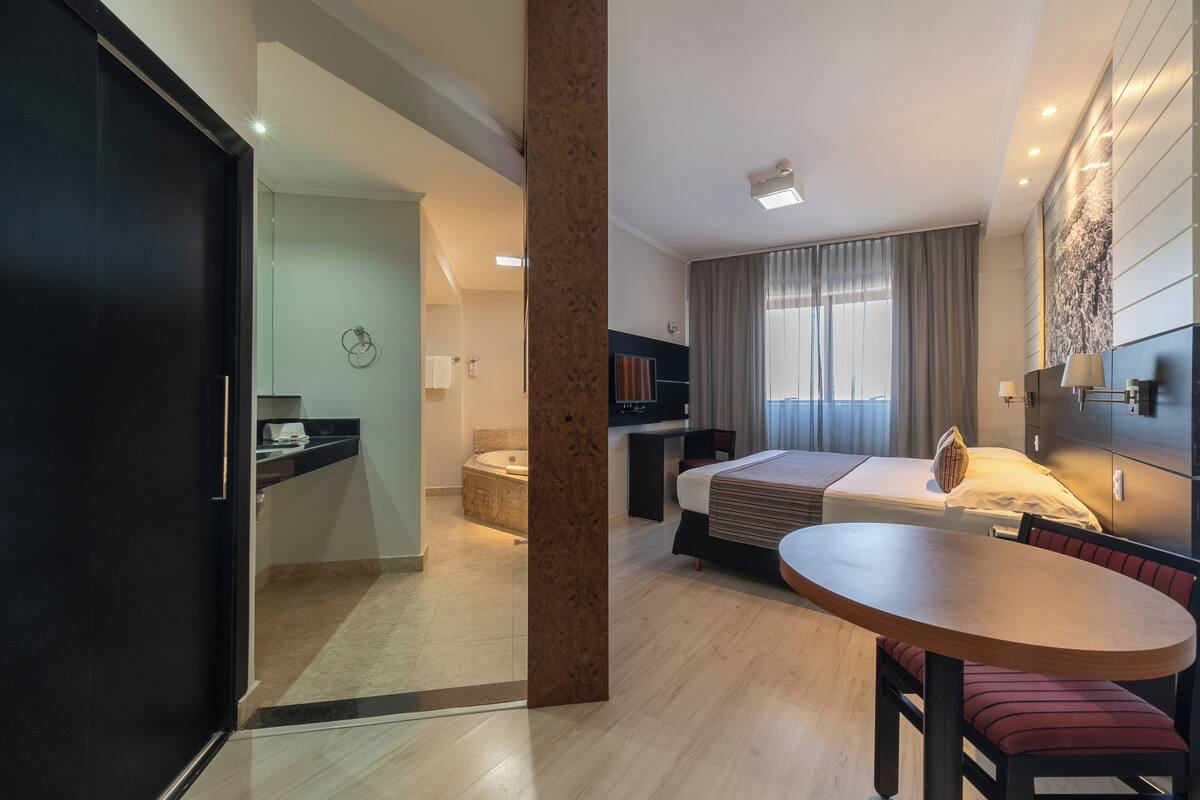 Quarto de hotel moderno com cama, mesa de jantar, pequena cozinha e banheiro.