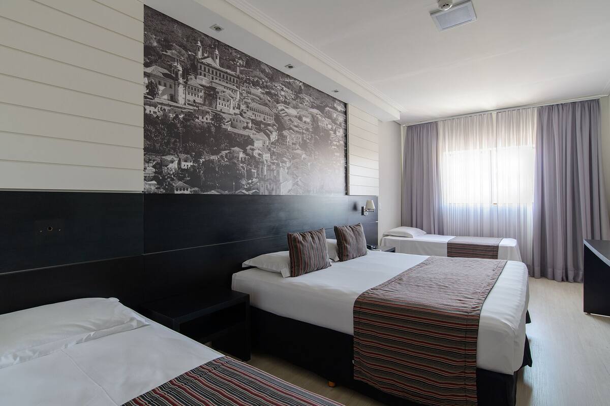 Quarto de hotel com uma cama grande, uma cama menor, uma escrivaninha e uma janela ampla com cortinas.