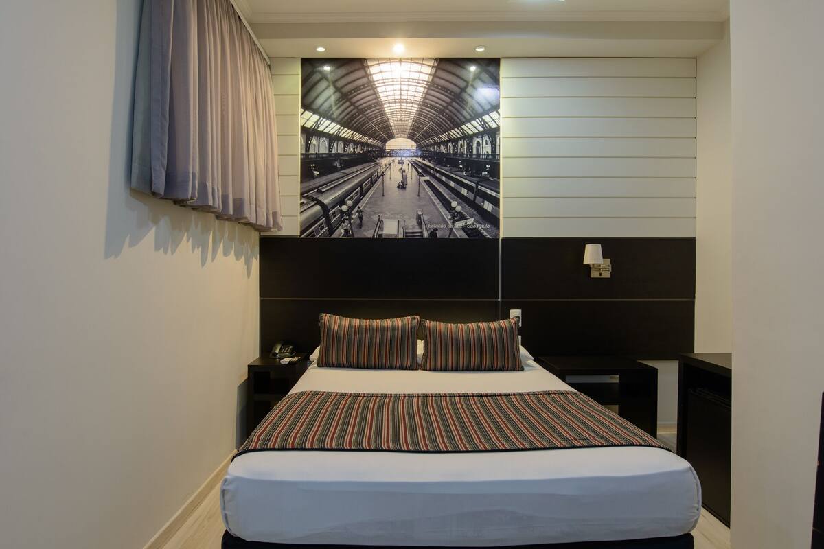 Quarto de hotel com uma cama grande, mesa de cabeceira com telefone, luminária na parede e vista para uma estação de trem.