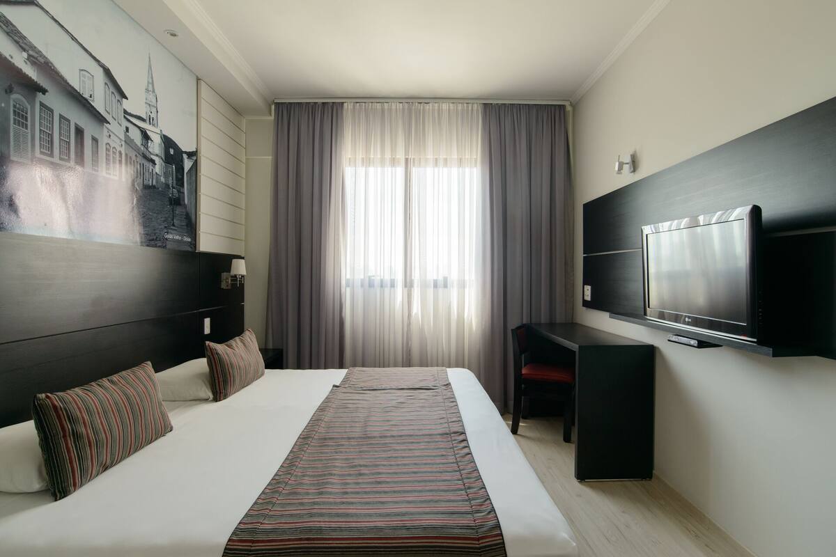 Quarto de hotel moderno com uma cama grande, uma TV de tela plana na parede e um quadro com vista para a cidade.