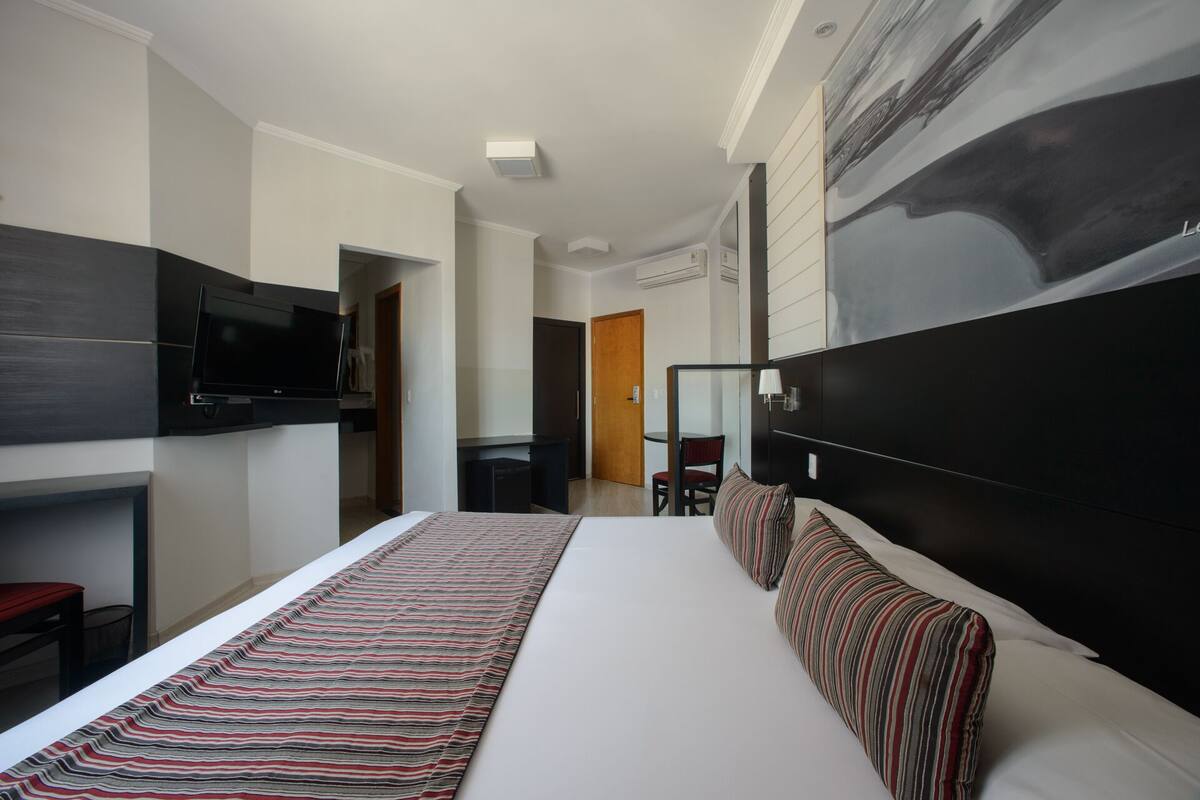 Quarto de hotel moderno com uma cama grande, TV de tela plana, escrivaninha e área de refeições.