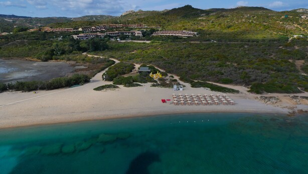 Vista aérea de um resort costeiro com uma praia de areia, águas turquesa cristalinas e um grande complexo hoteleiro ao fundo.