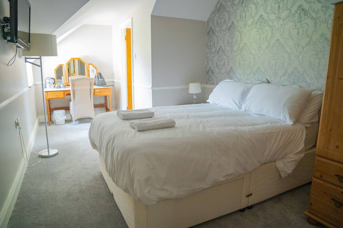 Quarto de hotel com uma cama grande, uma escrivaninha com cadeira, um espelho e uma cômoda.
