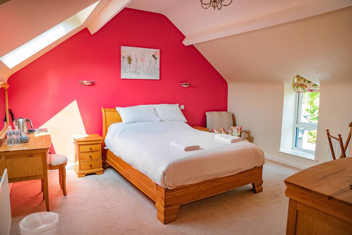 Quarto com cama de madeira, escrivaninha, cadeira, mesa de cabeceira e janela com vista para o verde.