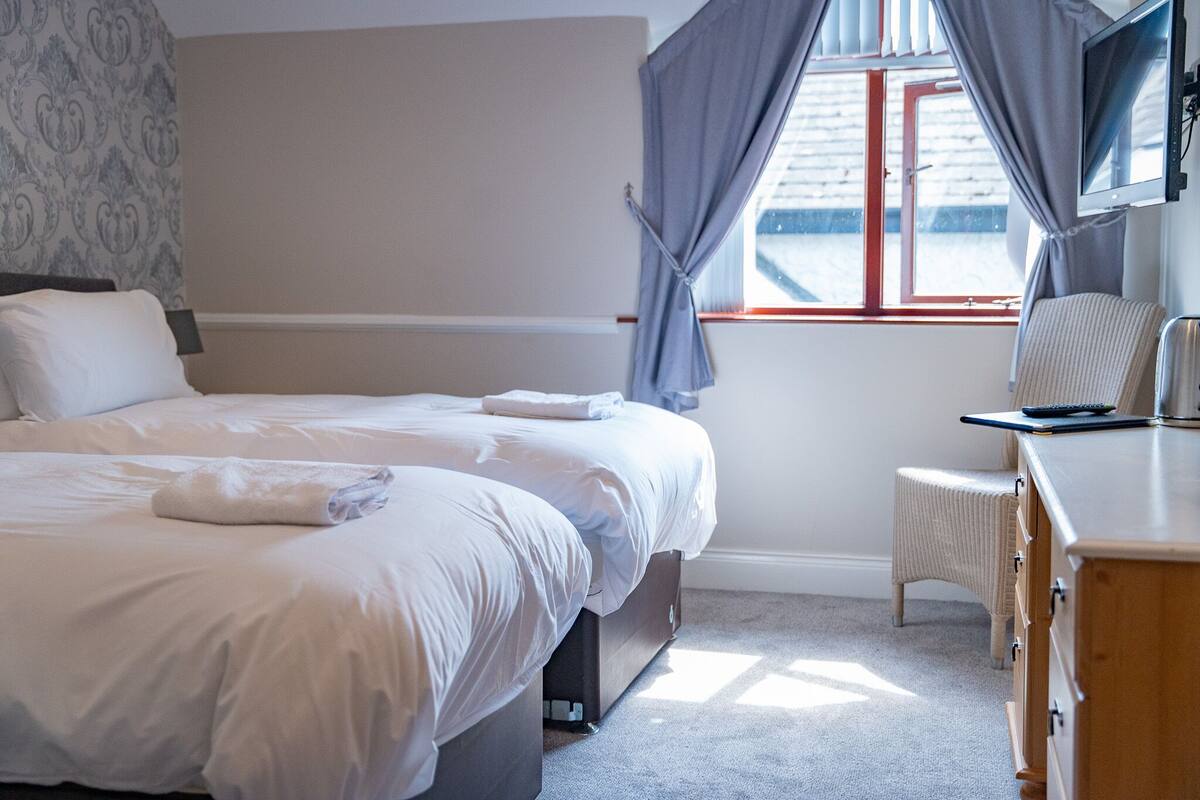 Cama bem arrumada com lençóis brancos, uma pequena mesa de cabeceira com abajur, uma cadeira e uma janela com cortinas.