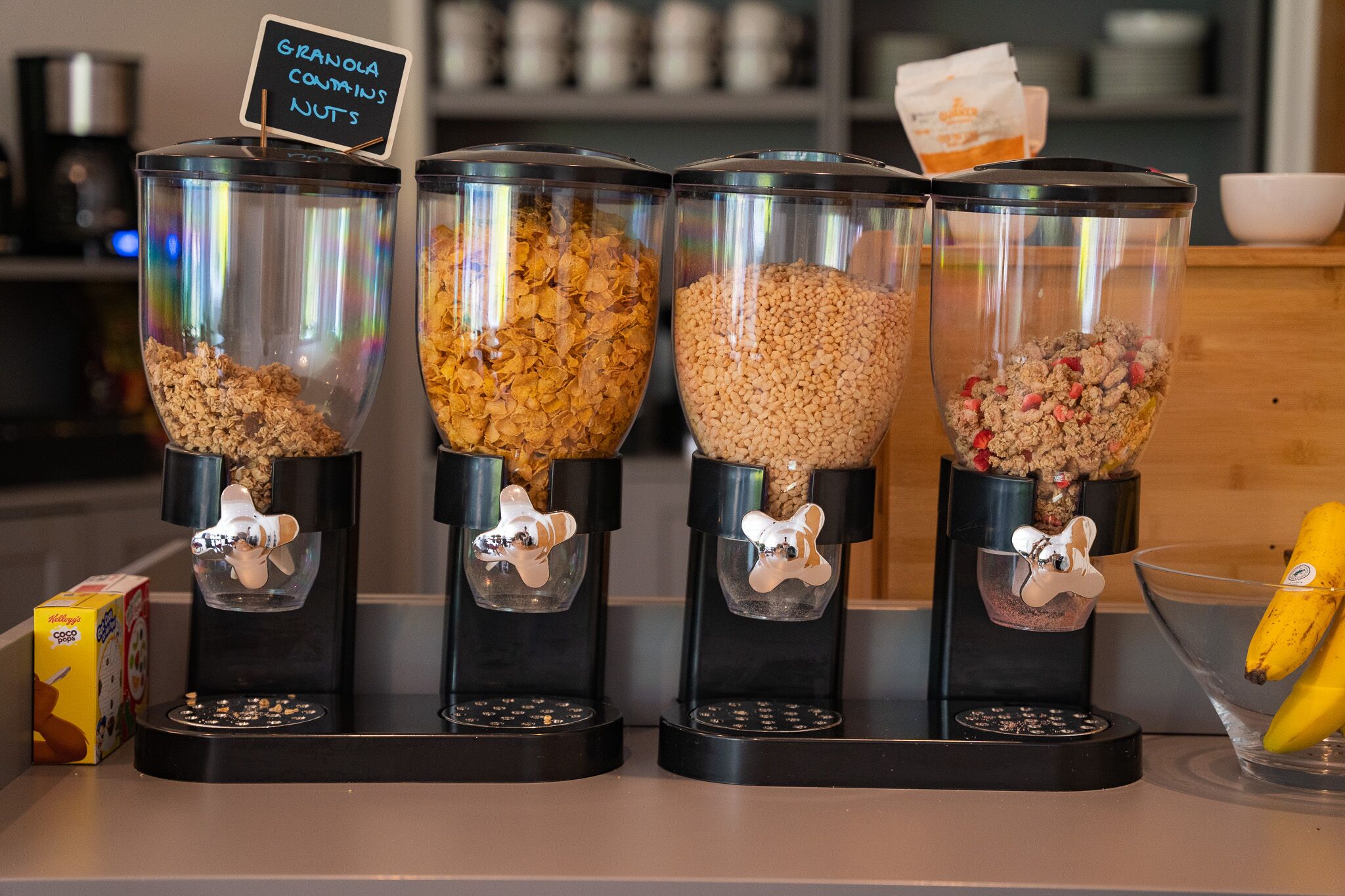 Quatro dispensadores com opções variadas de granola e frutos secos.