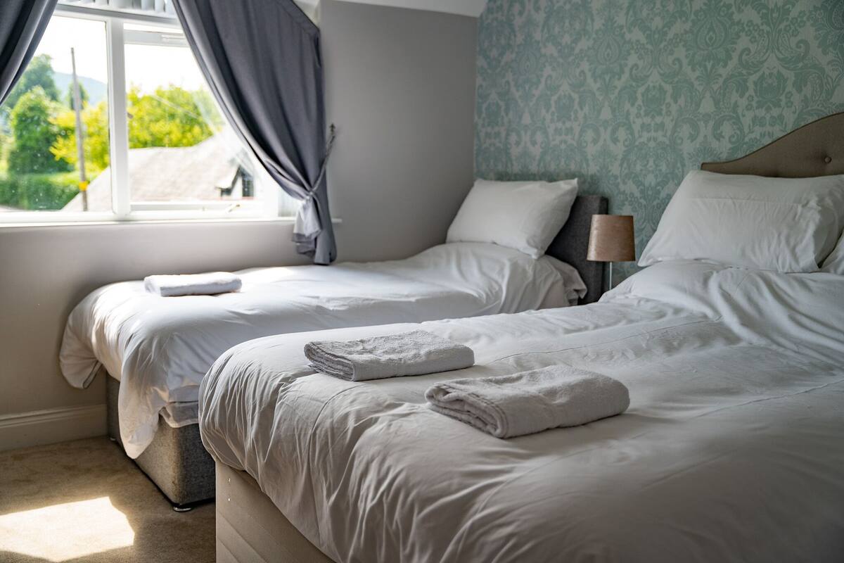 Quarto de hotel com duas camas, uma janela com cortinas, uma luminária e parede estampada.