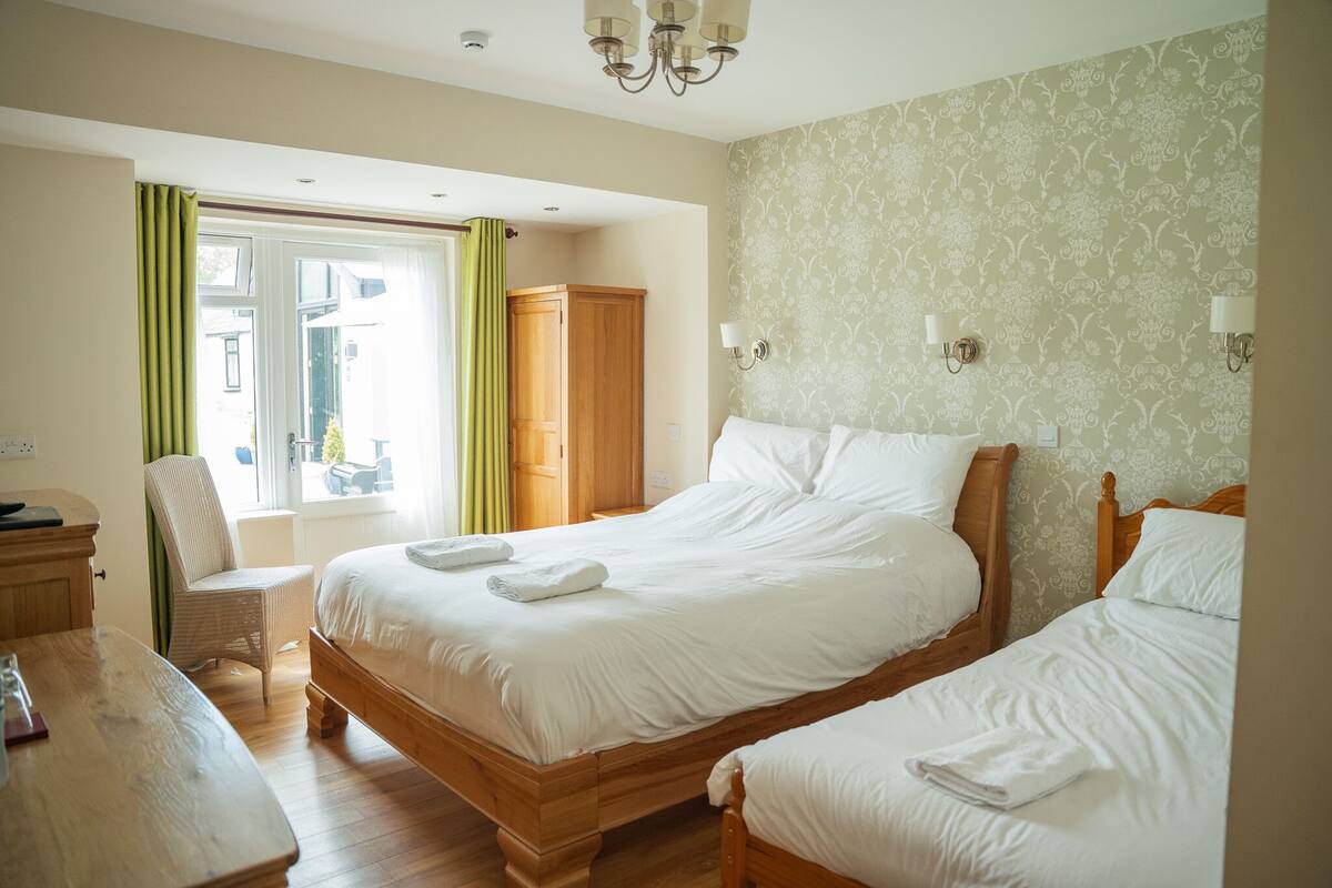 Quarto de hotel com duas camas, uma escrivaninha de madeira, uma cadeira, um guarda-roupa e uma janela com cortinas.