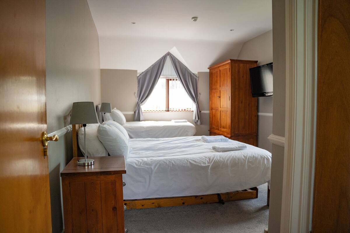 Quarto com duas camas, um guarda-roupa de madeira, uma televisão na parede e uma janela com cortinas.
