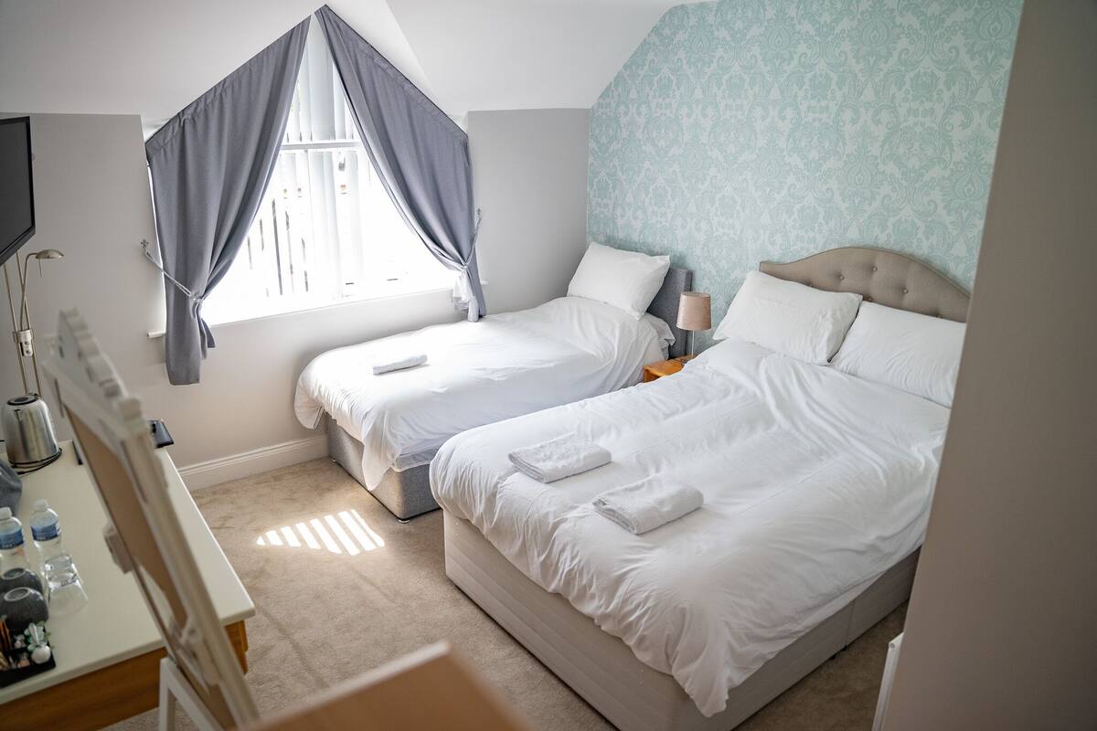 Quarto de hotel com duas camas, uma escrivaninha com abajur e uma janela com cortinas.
