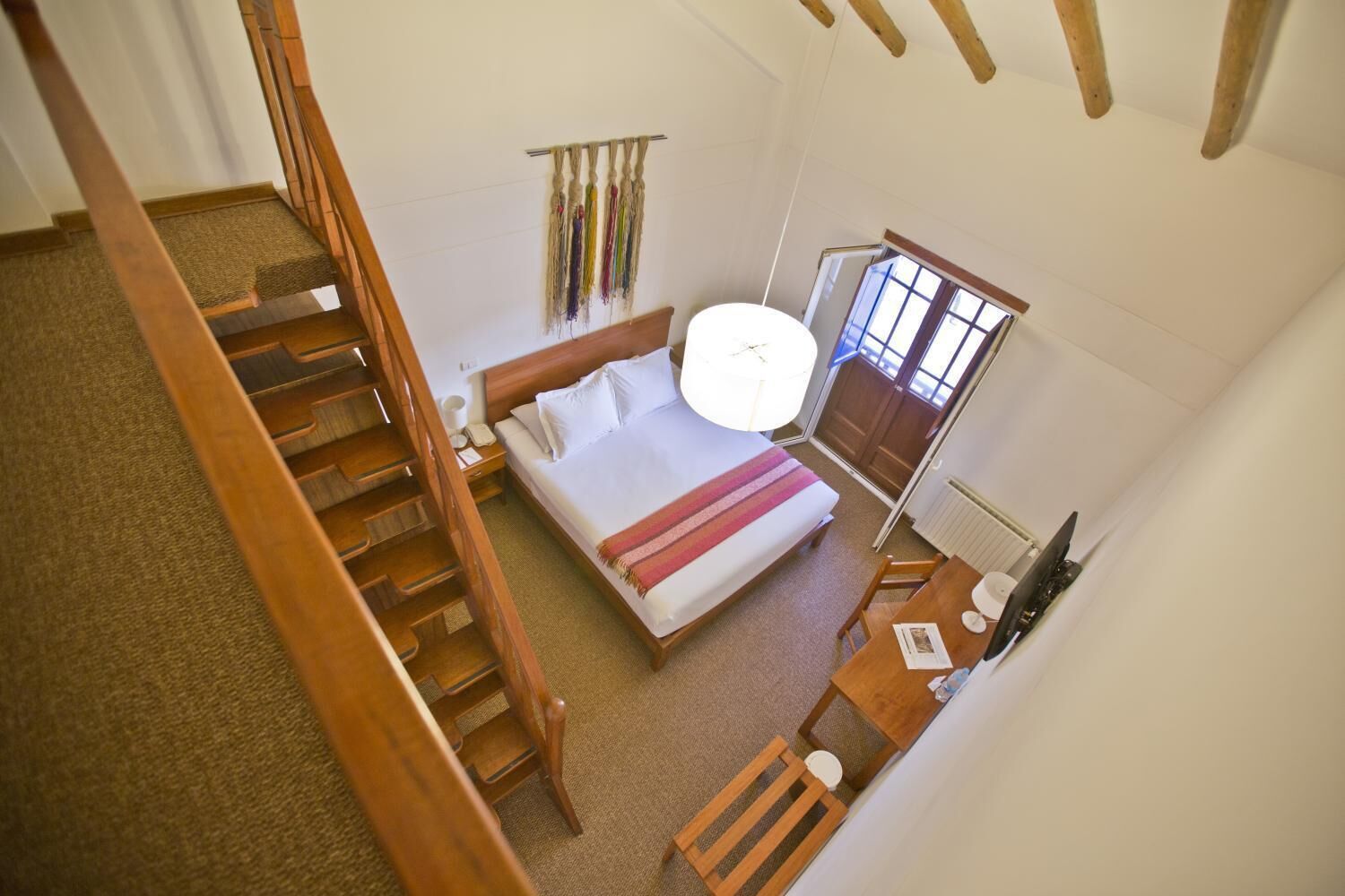 Quarto com cama, escrivaninha, cadeira, abajur e janela.