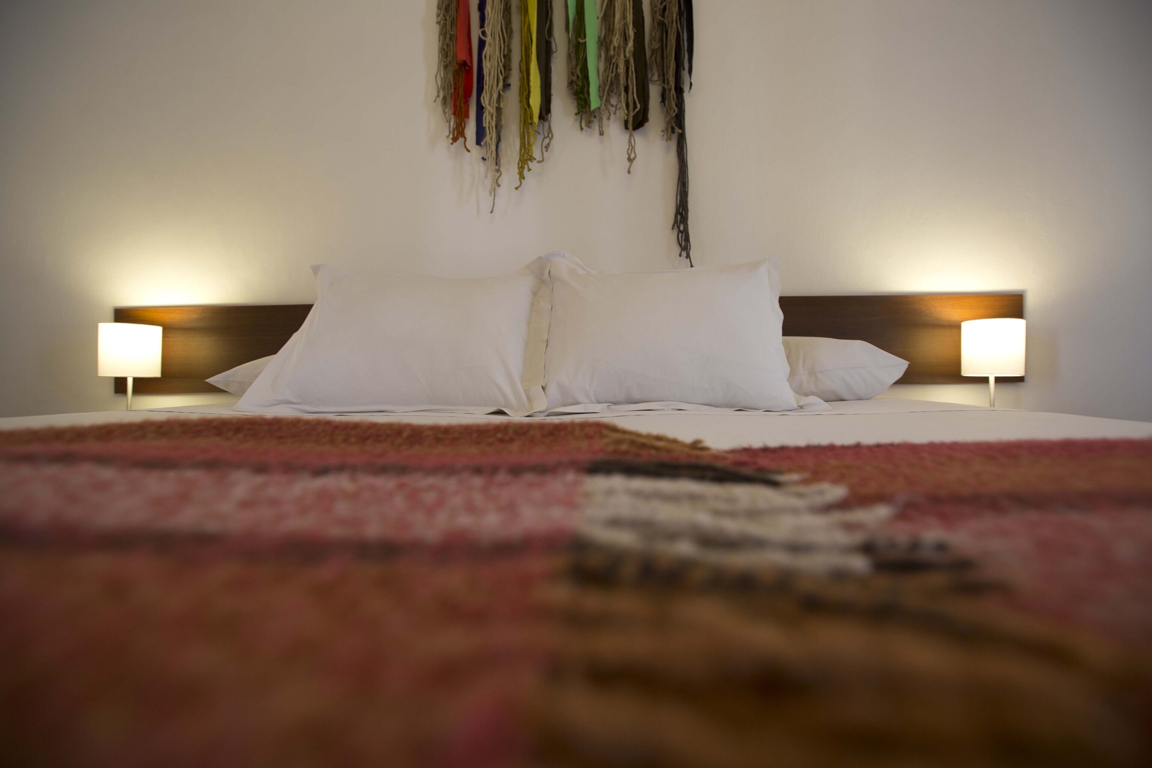 Uma cama com lençóis brancos, duas luminárias de cabeceira e uma tapeçaria colorida pendurada na parede.