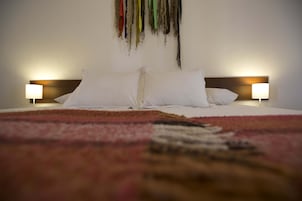 Uma cama com lençóis brancos, duas luminárias de cabeceira e uma tapeçaria colorida pendurada na parede.