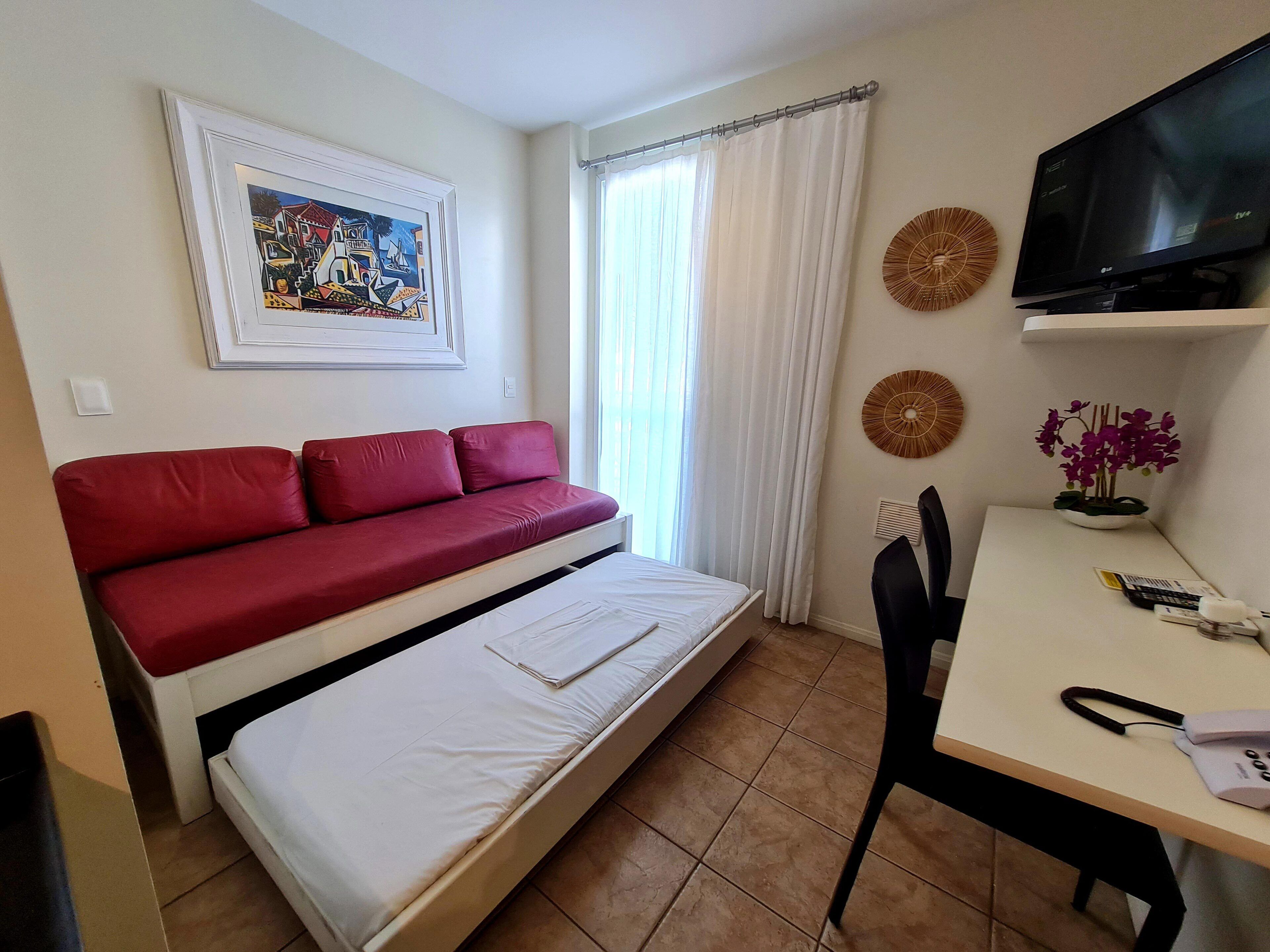 Quarto compacto com um sofá vermelho, uma cama pequena, uma escrivaninha com cadeira, televisão e um quadro na parede.