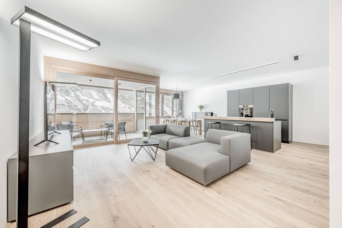 Sala de estar moderna com um sofá seccional cinza, uma mesa de centro e um espaço de jantar com mesa e cadeiras. Há também uma cozinha com armários cinzas e bancada.