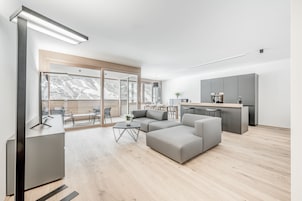 Sala de estar moderna com um sofá seccional cinza, uma mesa de centro e um espaço de jantar com mesa e cadeiras. Há também uma cozinha com armários cinzas e bancada.