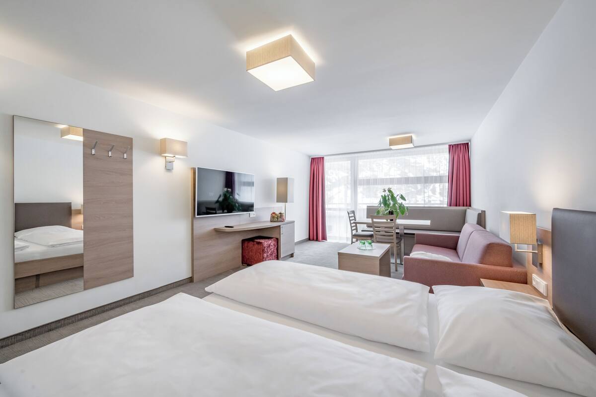 Quarto de hotel moderno com cama grande, escrivaninha, sofá, televisão e uma janela ampla com cortinas.
