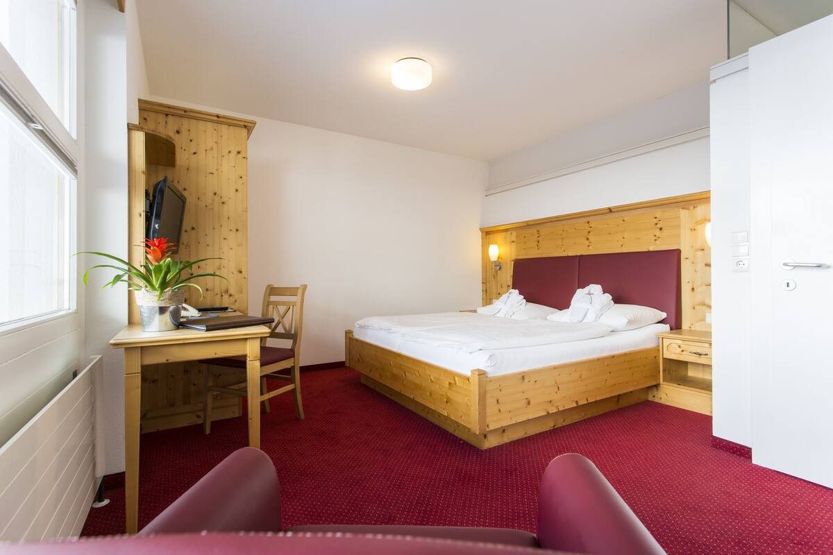 Quarto de hotel com cama de madeira, escrivaninha com cadeira, pequena mesa com planta e chão coberto por um tapete vermelho.