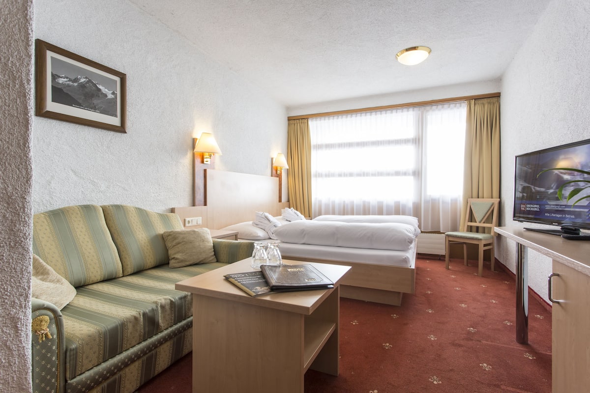 Quarto de hotel com sofá, cama, escrivaninha com um laptop, televisão e uma moldura com uma imagem na parede.
