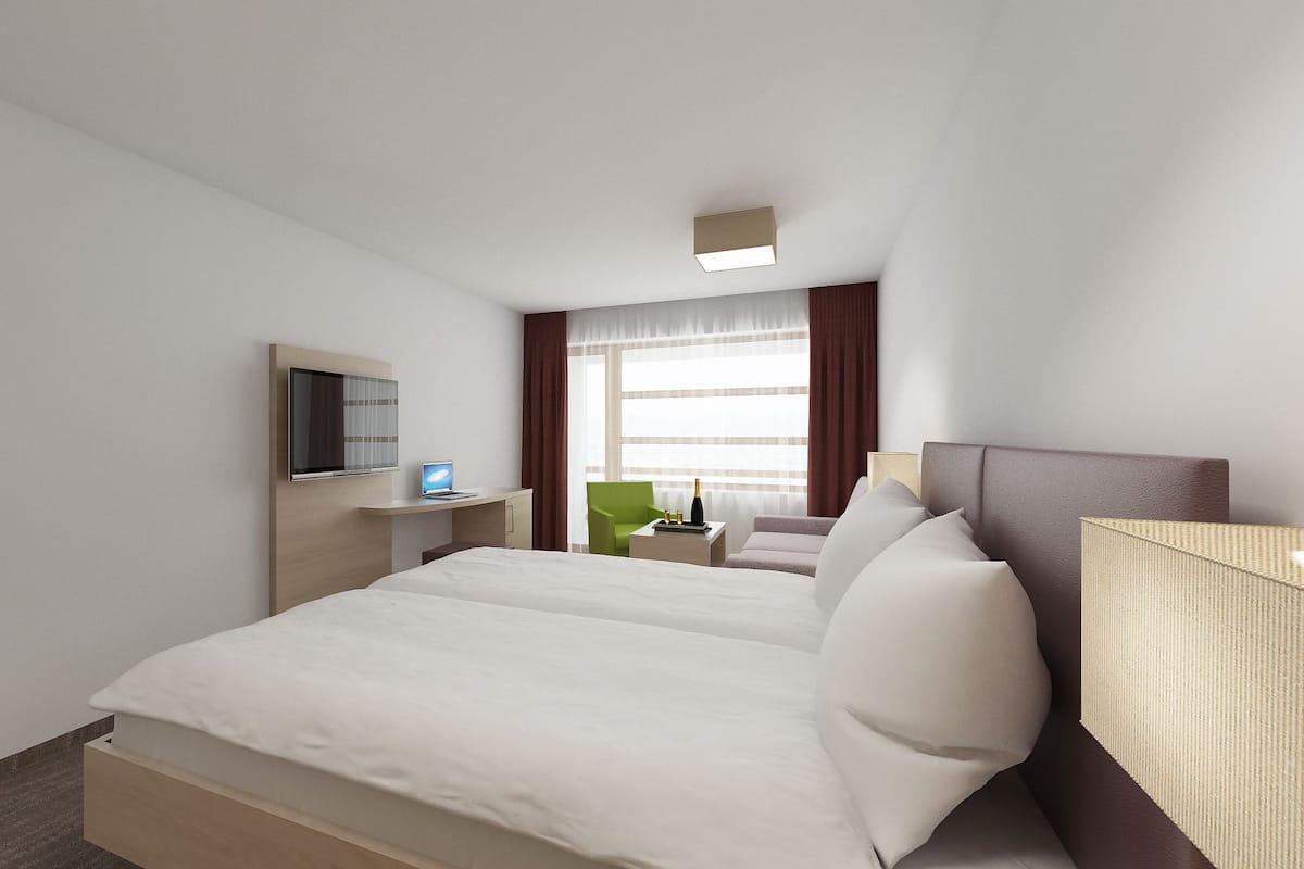 Quarto de hotel com uma cama, uma mesa com um computador, uma cadeira e uma janela com cortinas.