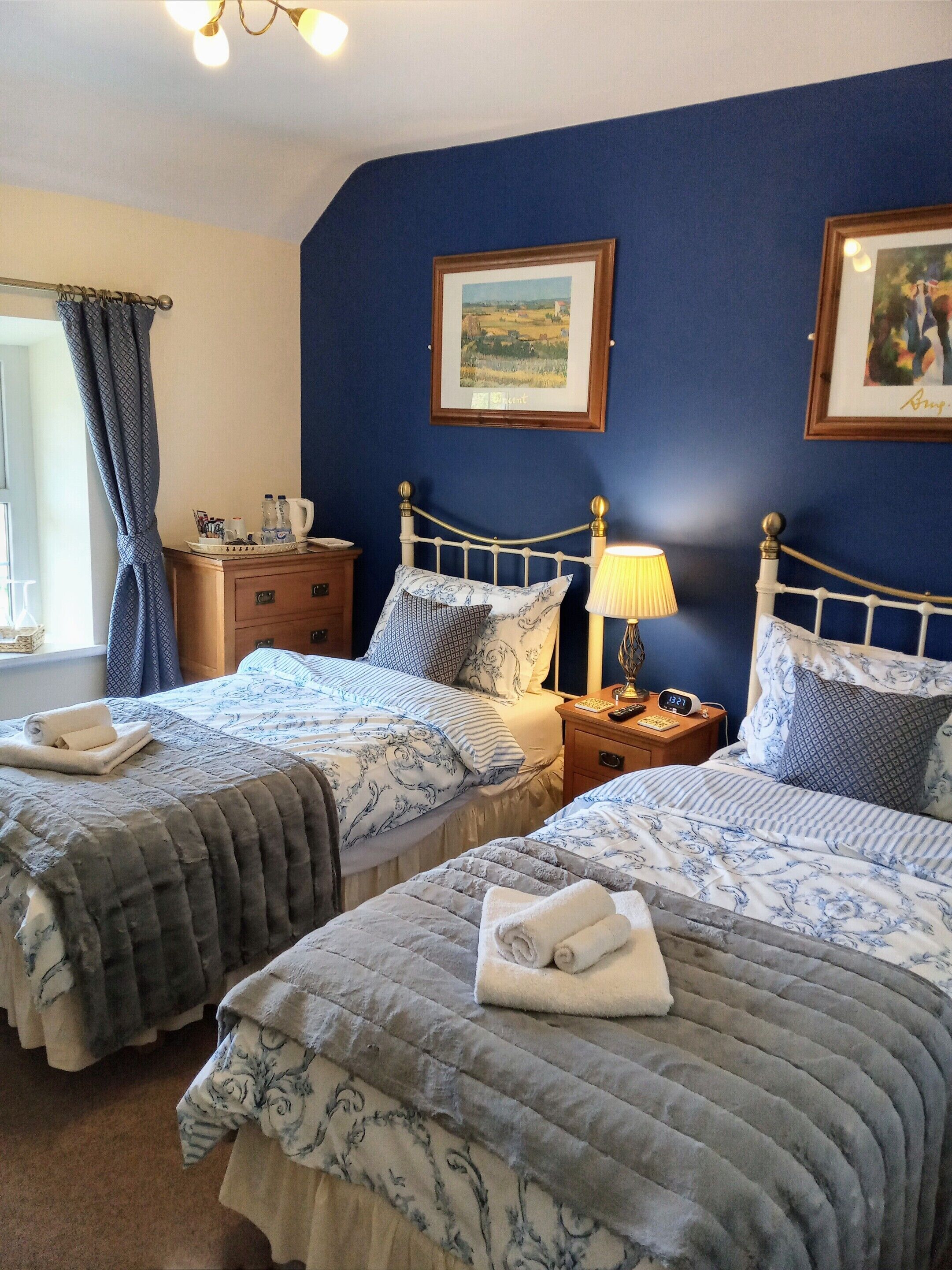 Quarto de hotel com duas camas, mesa de cabeceira com abajur, cômoda e janela com cortinas.