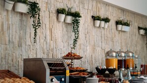 Buffet de café da manhã com diversos pães, uma máquina de café e dispensadores de suco.