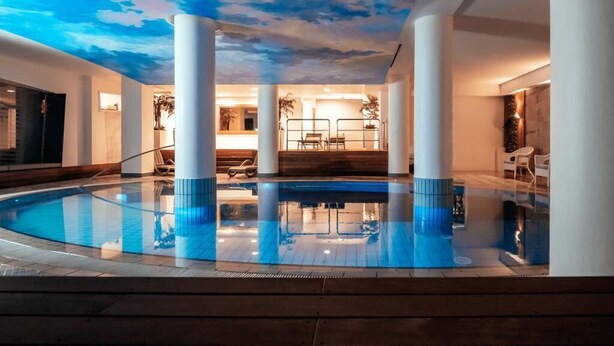 Piscina interna com teto que simula um céu com nuvens, cercada por pilares brancos e um deck de madeira.