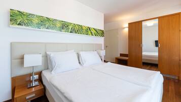 Quarto de hotel com uma cama grande, uma mesa de cabeceira com luminária, guarda-roupa de madeira e uma obra de arte na parede.