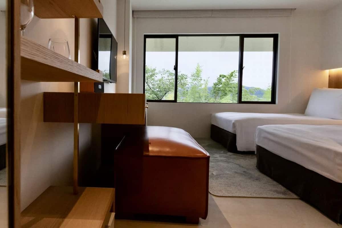 Quarto de hotel com duas camas, um banco de madeira, uma TV e uma janela grande com vista.