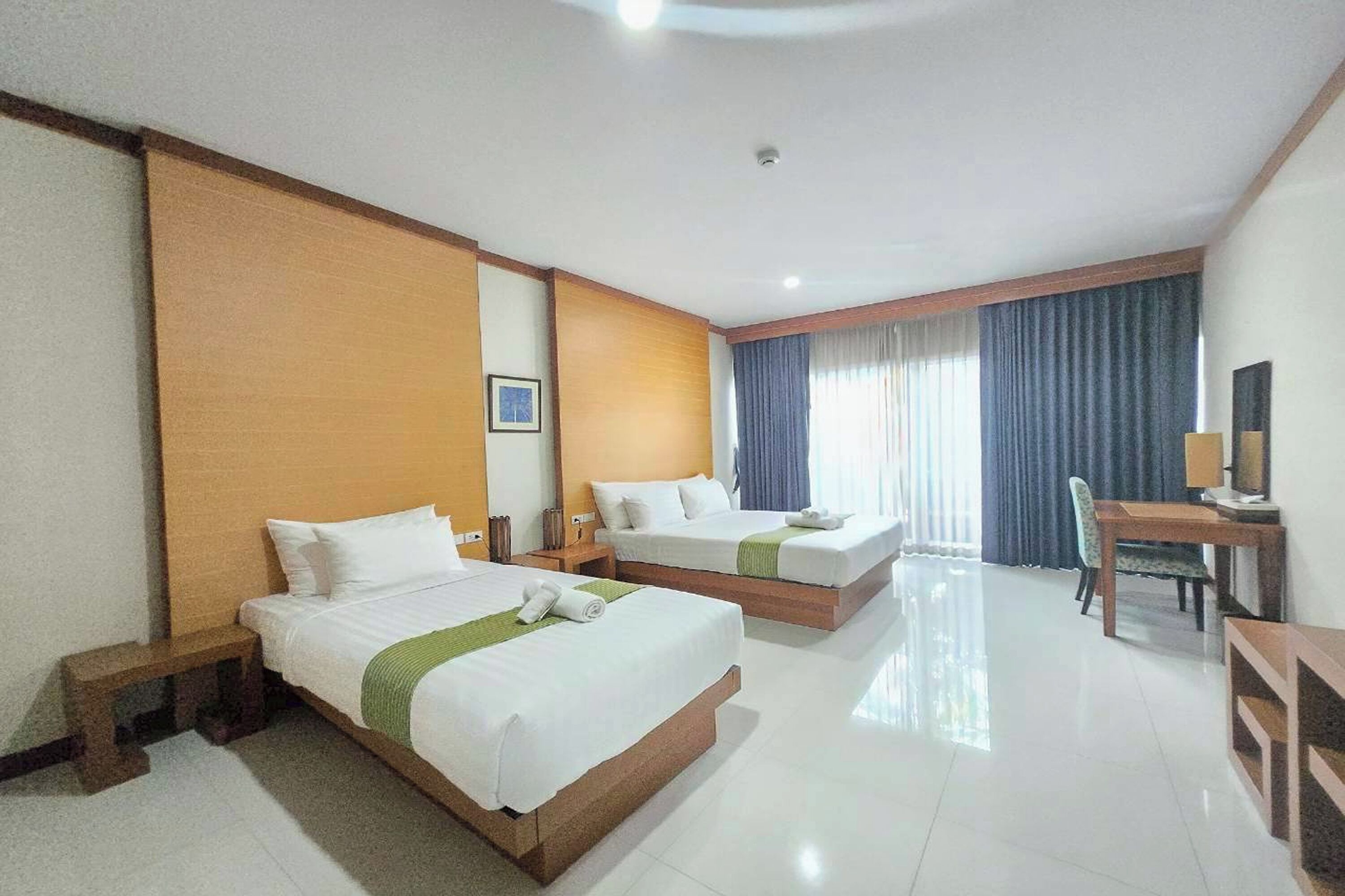Quarto de hotel com duas camas, uma escrivaninha, uma cadeira e uma janela grande com cortinas.