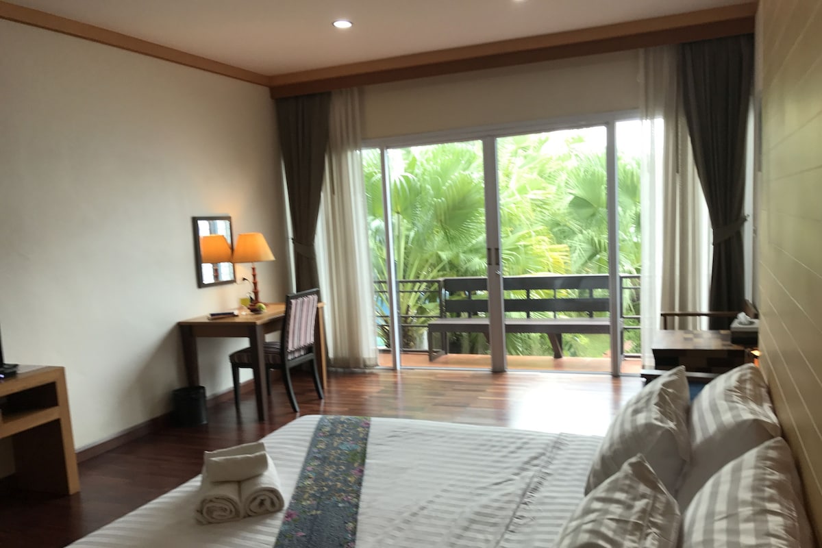 Quarto de hotel com cama, escrivaninha, cadeira, mesa e varanda com vista para o verde.