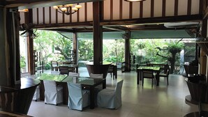 Um espaço de refeições interno com mesas e cadeiras de vidro, vigas de madeira e amplas janelas com vista para um jardim.