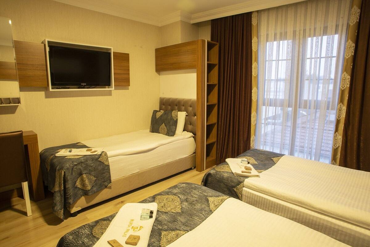Quarto de hotel com duas camas, televisão na parede, guarda-roupa de madeira e janela com cortinas.