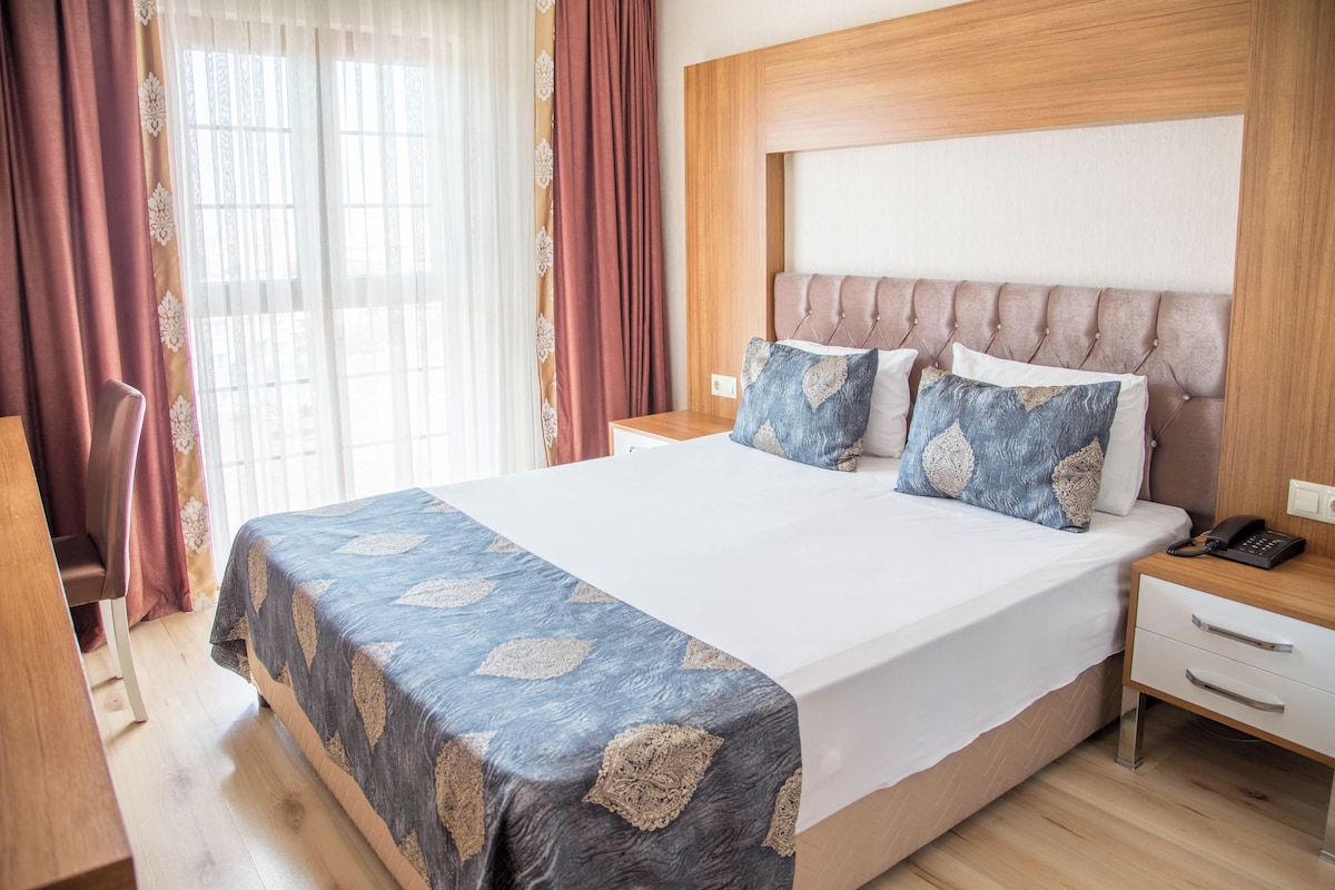 Quarto de hotel com uma cama grande, cabeceira de madeira, mesa de cabeceira com telefone e escrivaninha com cadeira.