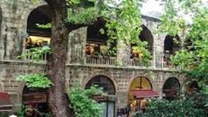 Um edifício de pedra com vários andares, janelas em arco e varandas, rodeado por vegetação exuberante.